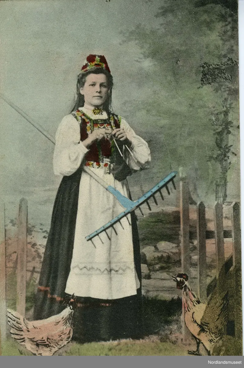 Postkort som viser kvinne i bunad. Hun står i en gjerdeåpning og holder ...