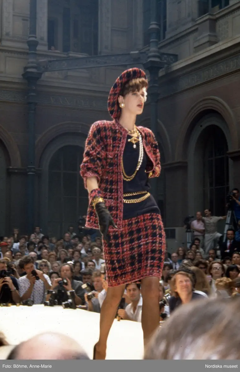 Modevisning. Modell i rödrutig dräkt, hatt och gulddetaljer. Från ...