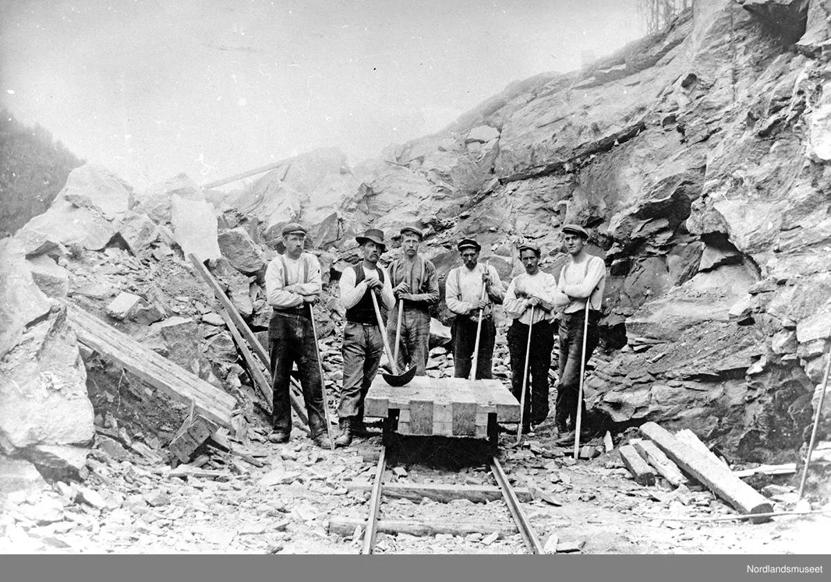 Arbeidere med spett og slegge. Fjellmasse bak dem, jernbaneskinner i ...