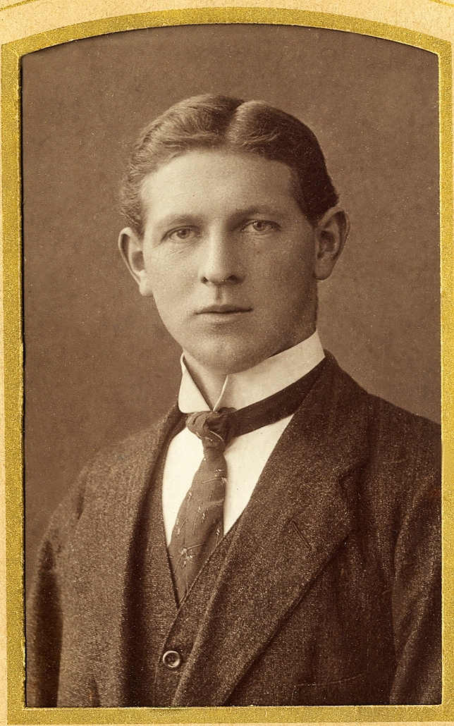 En man i mörk kostym med väst, stärkkrage och slips. Under fotot text ...