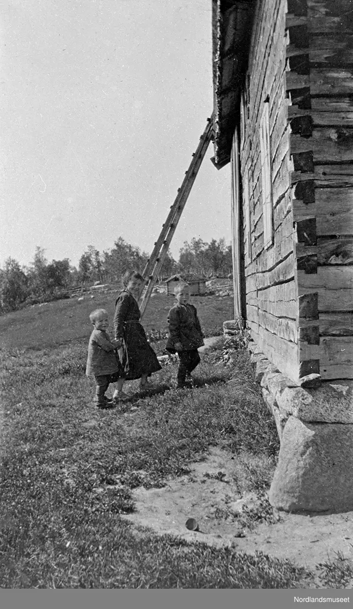 Tre barn som står ved siden av en husvegg. En stige er reist opp mot ...